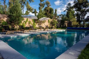 Piscina com espreguiçadeiras e guarda-sóis, cercada por árvores e um muro de pedra.
