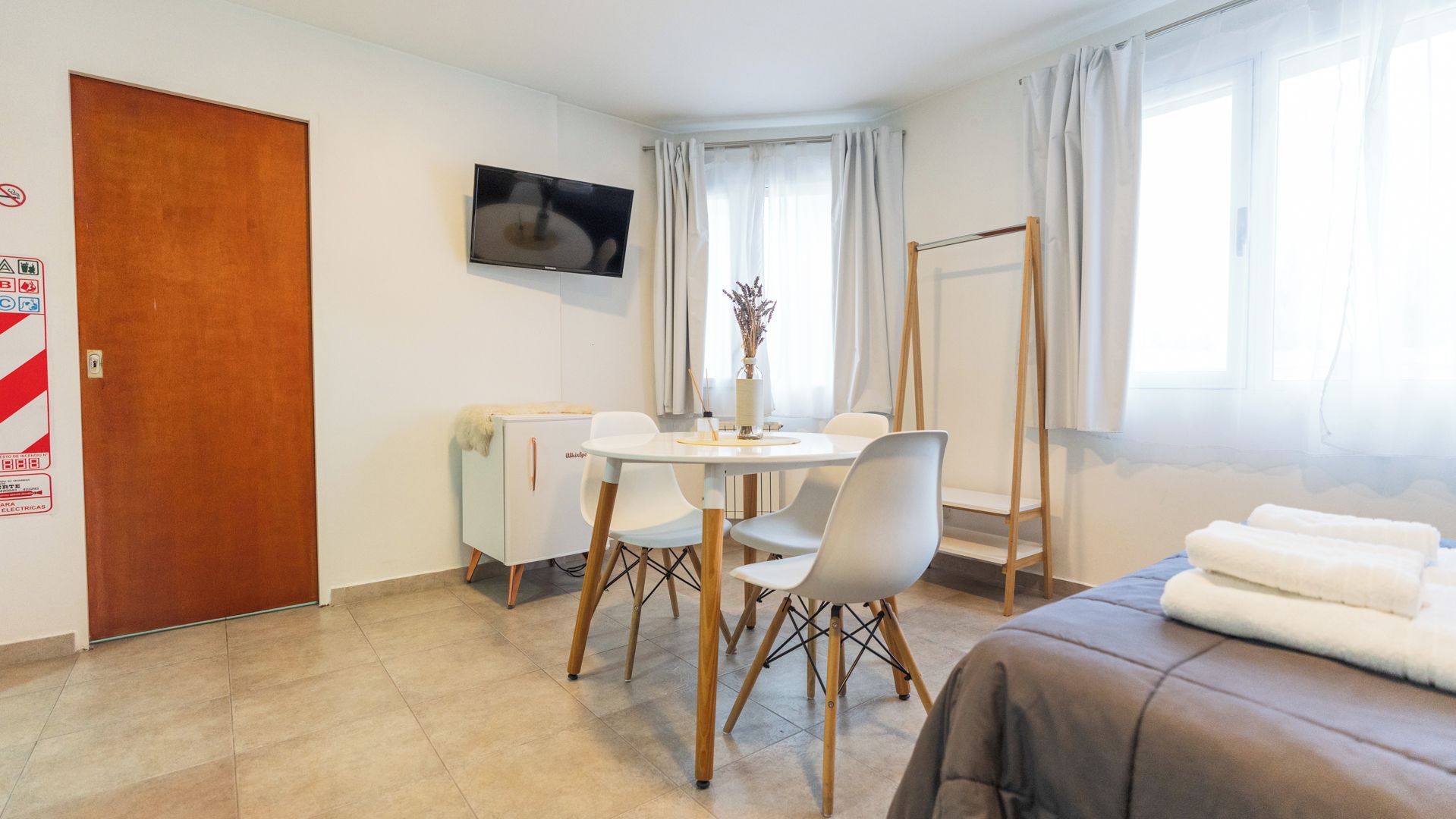 Quarto de hotel com cama, mesa de jantar com cadeiras, televisão na parede e janela com cortinas.