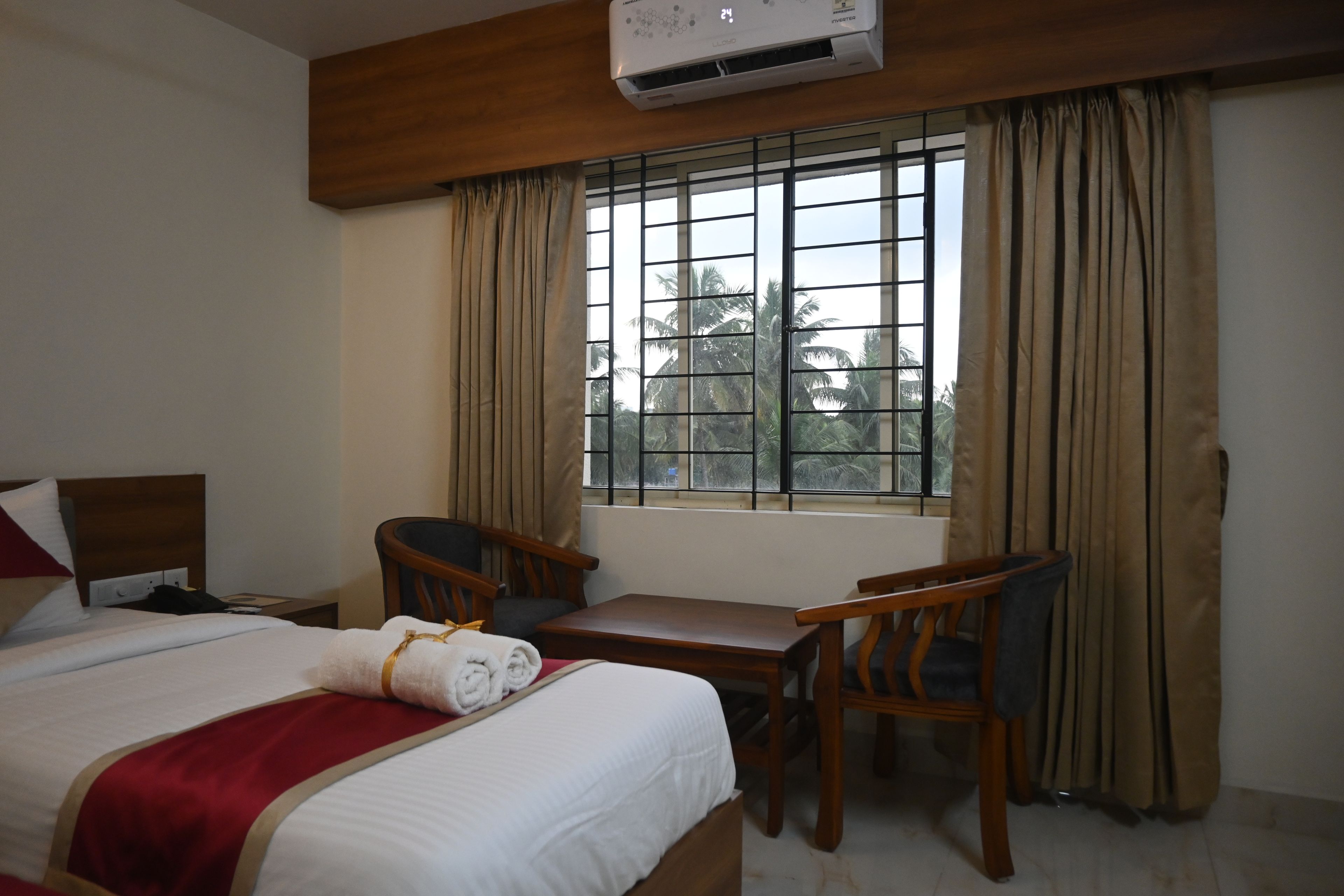 Quarto de hotel com cama, mesa, duas cadeiras, janela com cortinas e aparelho de ar condicionado.