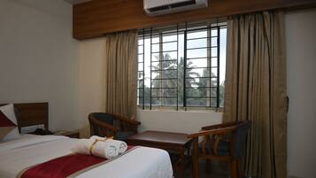 Quarto de hotel com cama, mesa, duas cadeiras, janela com cortinas e aparelho de ar condicionado.