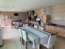 Uma sala de jantar com uma mesa comprida, cadeiras e uma cozinha com painel de azulejos e armários de madeira.