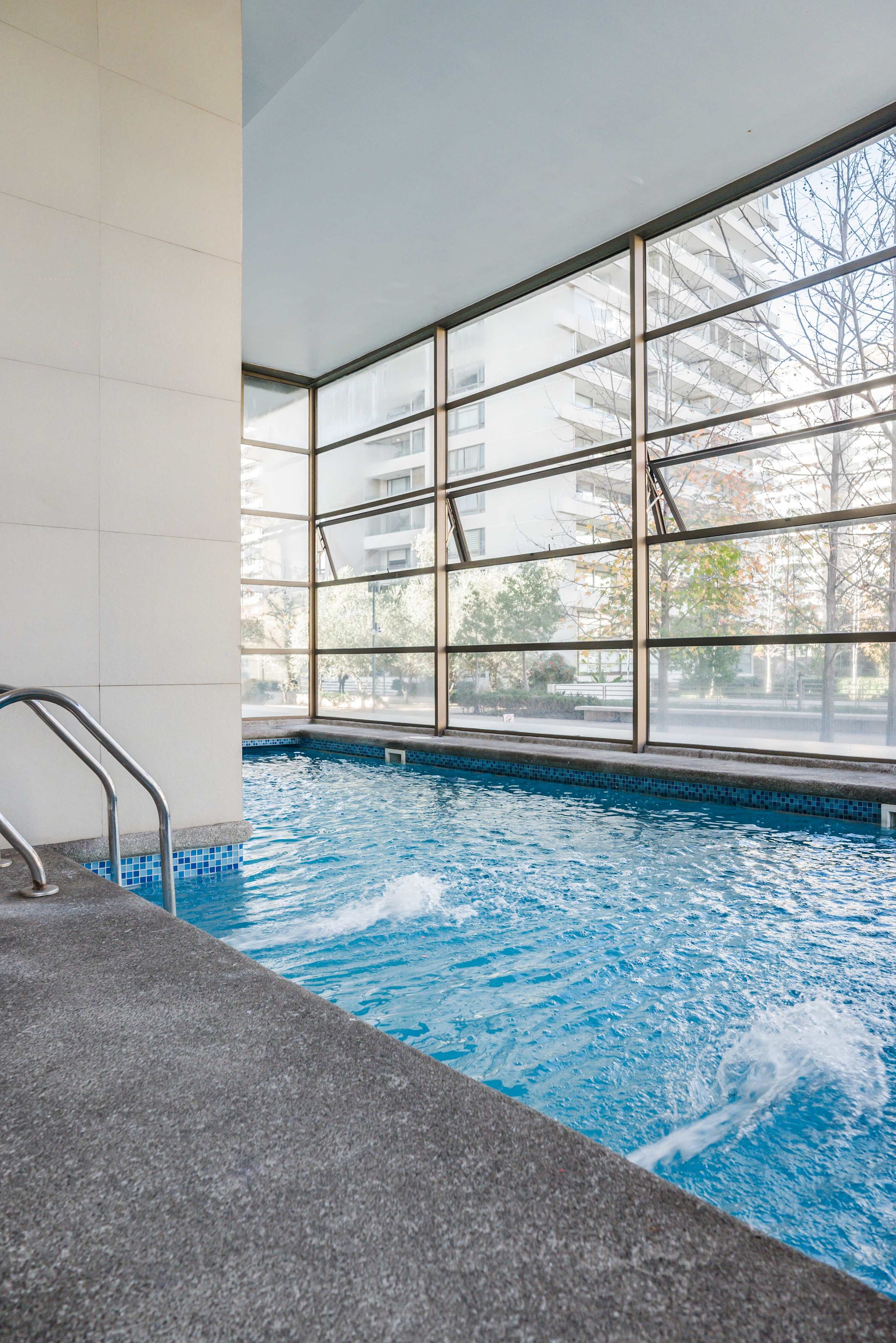 Piscina interior com paredes de vidro transparente, chão revestido com azulejos e escada de metal.