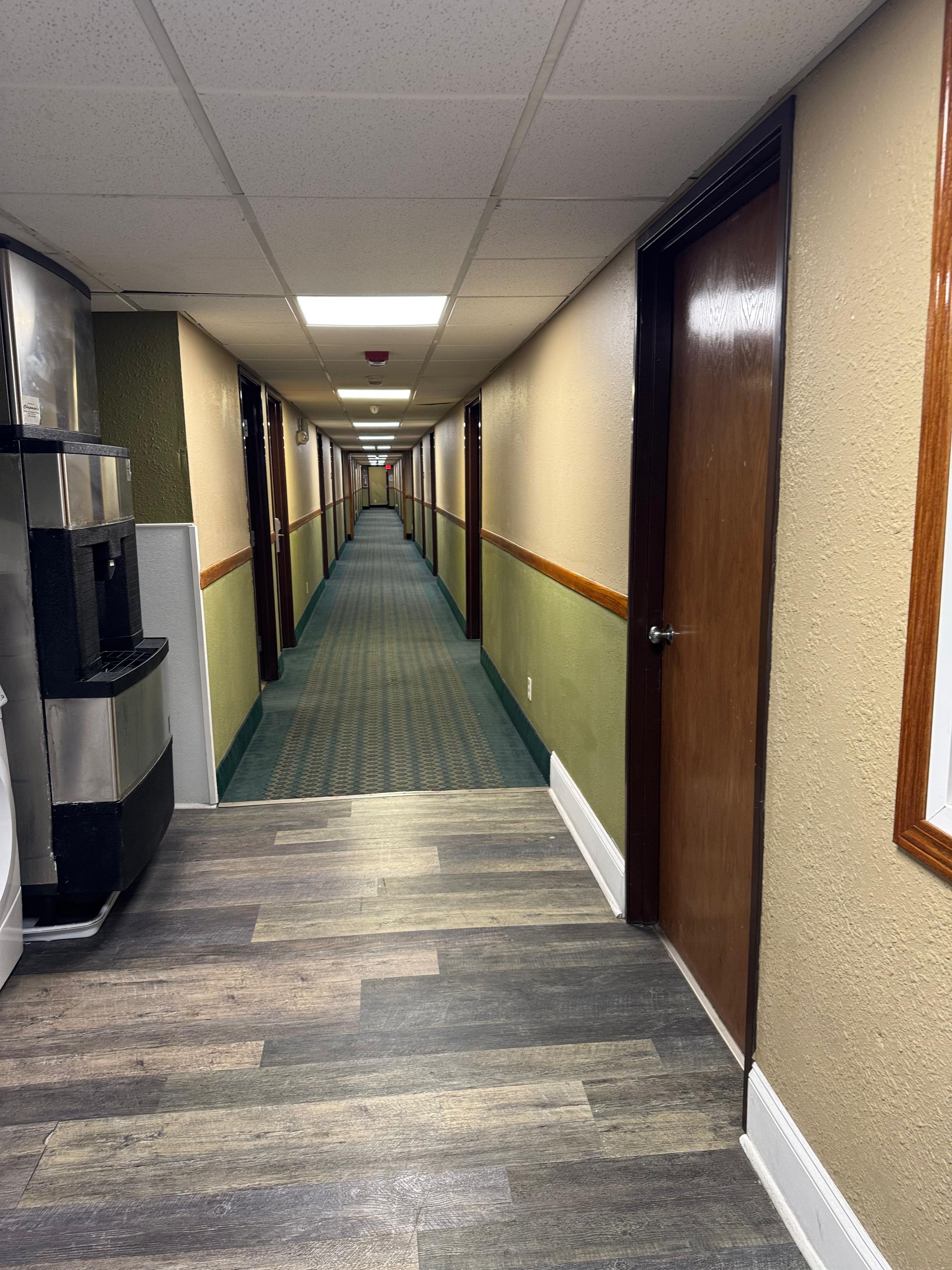 Corredor de hotel com um longo corredor, portas de madeira e uma área de kitchenette com uma máquina de café.