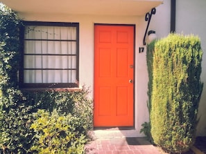 Uma casa com uma porta laranja vibrante, uma janela com padrão quadriculado e um alto cercado de sebe verde.