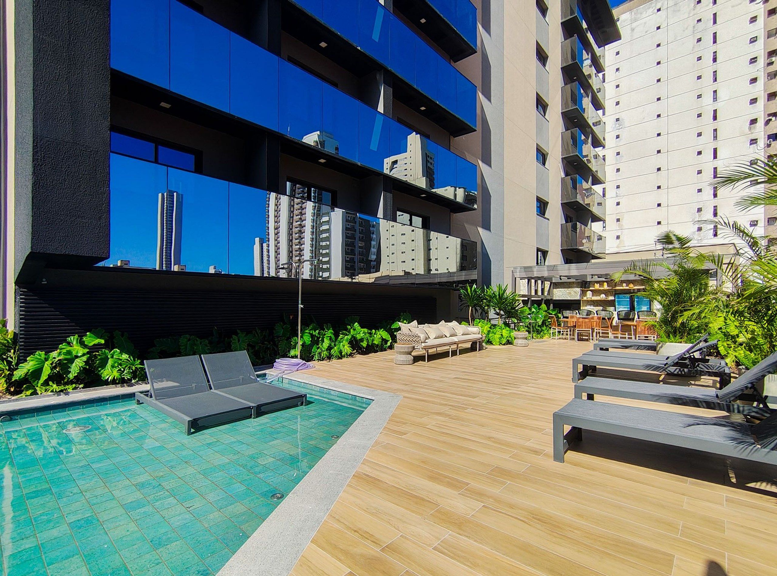 Área de piscina no terraço com espreguiçadeiras e um pequeno espaço de estar, cercada por vegetação e um edifício moderno com janelas de vidro refletivas.