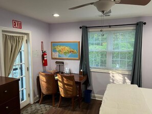 Quarto de hotel com cama, escrivaninha com cadeiras, mapa na parede, placa de saída e janela com cortinas.
