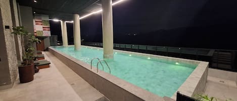 Piscina interna com água cristalina, cercada por colunas e plantas em vasos, sob um céu noturno escuro.