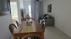 Uma sala de jantar com uma mesa de madeira, cadeiras e uma tigela decorativa. Um sofá e cortinas são visíveis ao fundo.