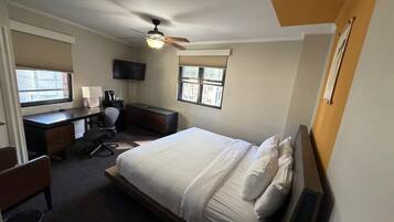 Quarto de hotel com cama, escrivaninha, cadeira, televisão e janelas.