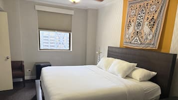 Quarto de hotel com uma cama grande, janela com persianas, uma cadeira, escrivaninha e tapeçaria decorativa na parede.
