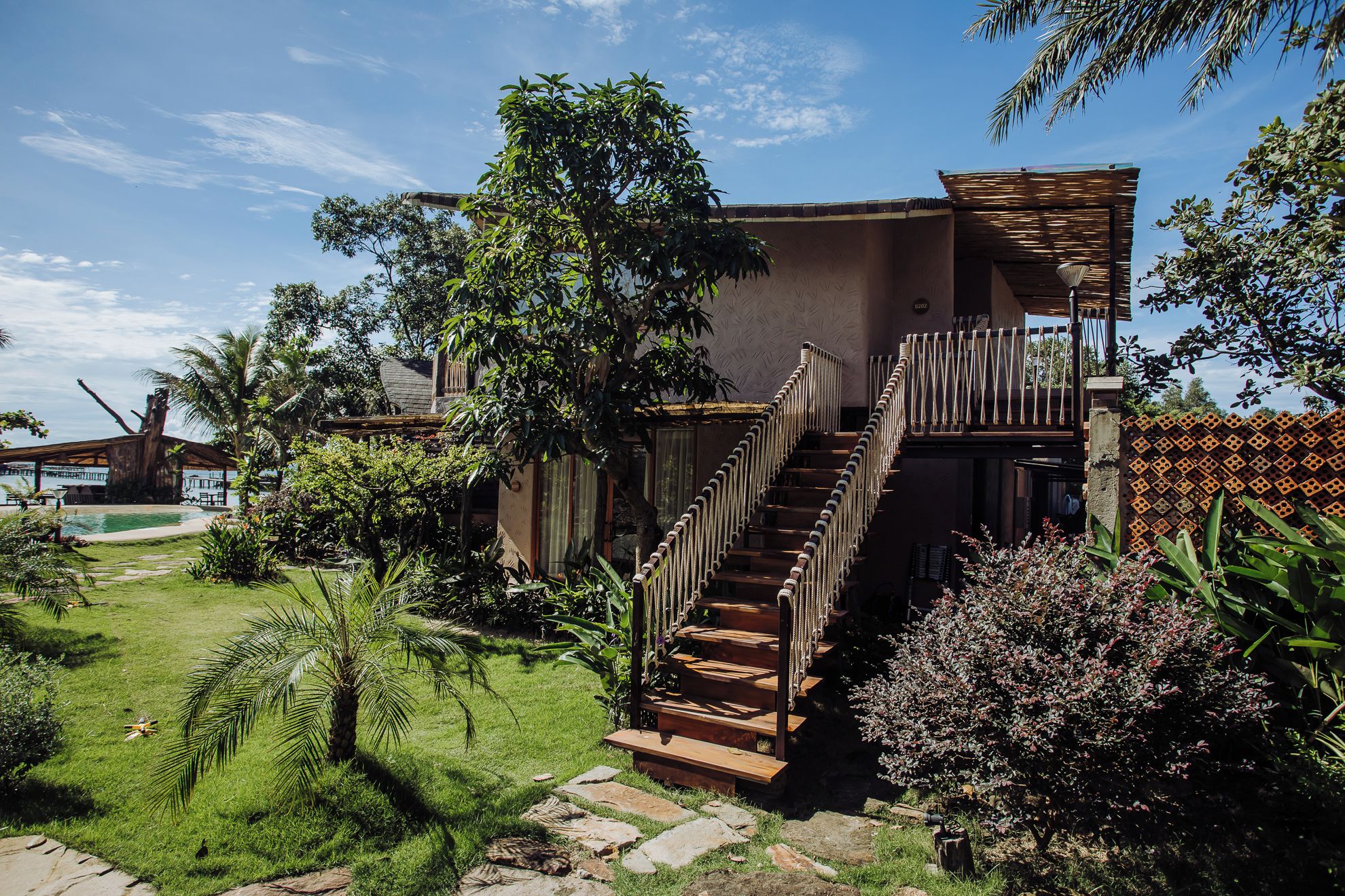 Um imóvel residencial com escada de madeira que dá para uma varanda, cercado por vegetação exuberante e uma piscina.
