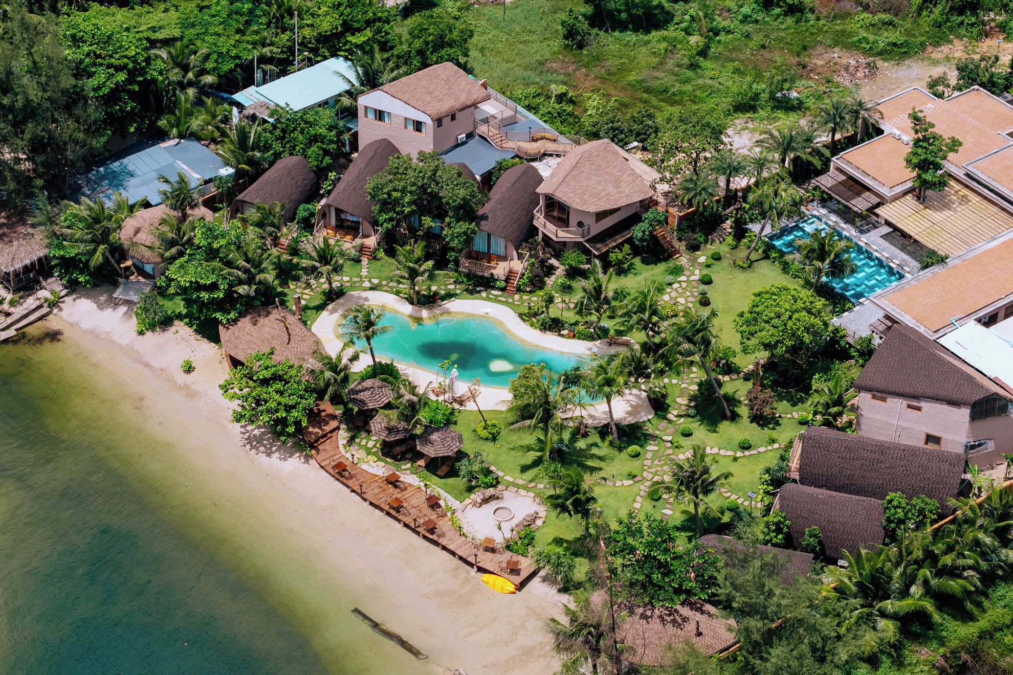 Vista aérea de um resort com piscina, praia e vários edifícios, tudo cercado por vegetação exuberante.