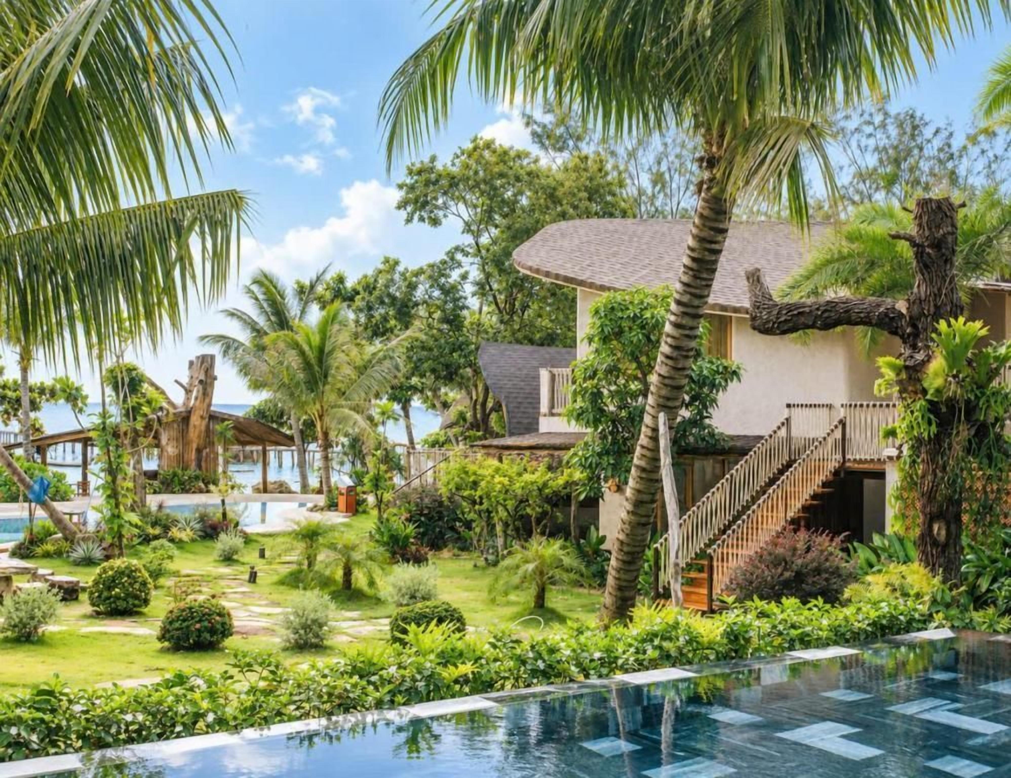 Vista da piscina em um resort, com céu claro, vegetação exuberante e um edifício com escadaria.