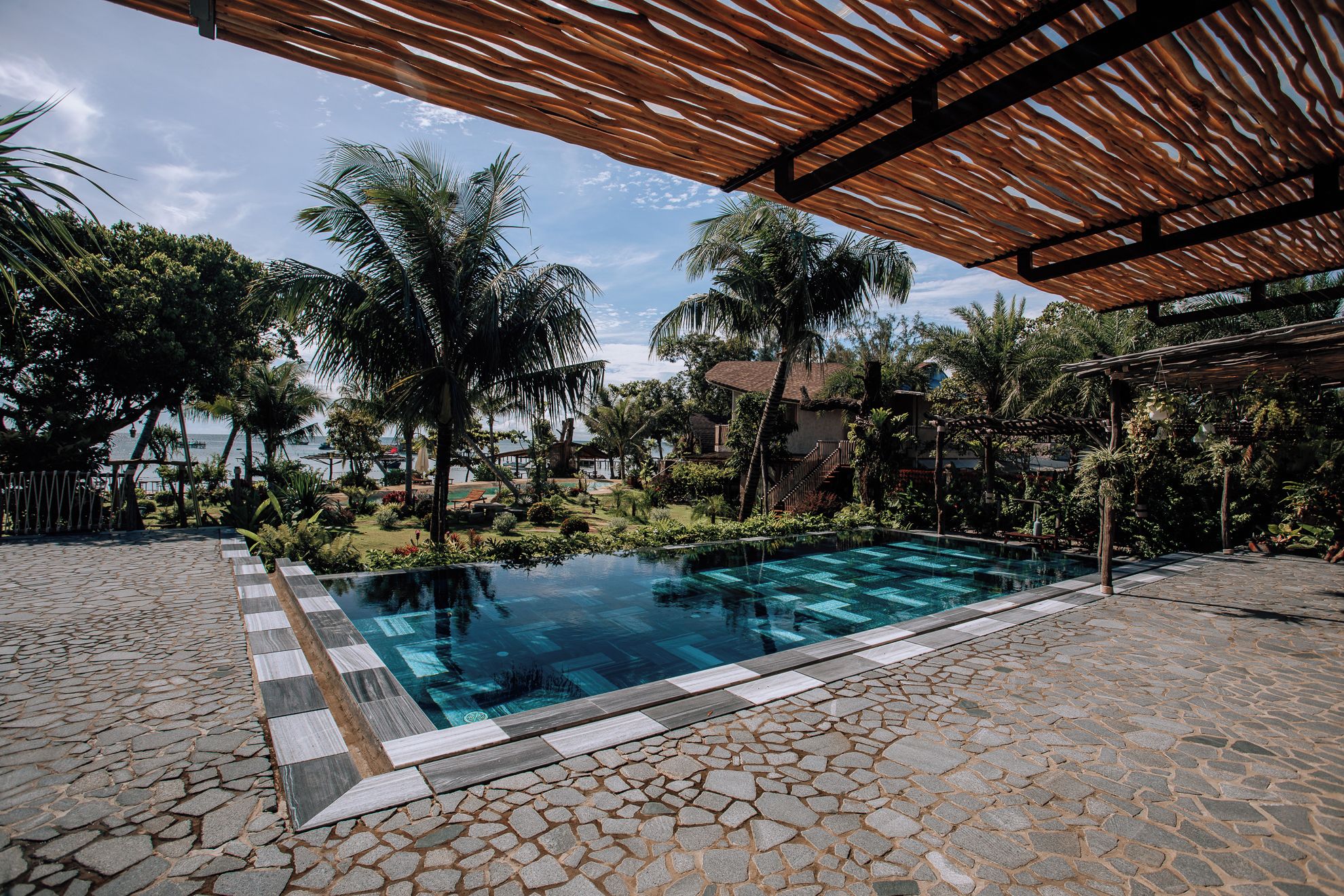 Piscina com borda de pedra e azulejos, rodeada por palmeiras e uma pérgula de madeira.