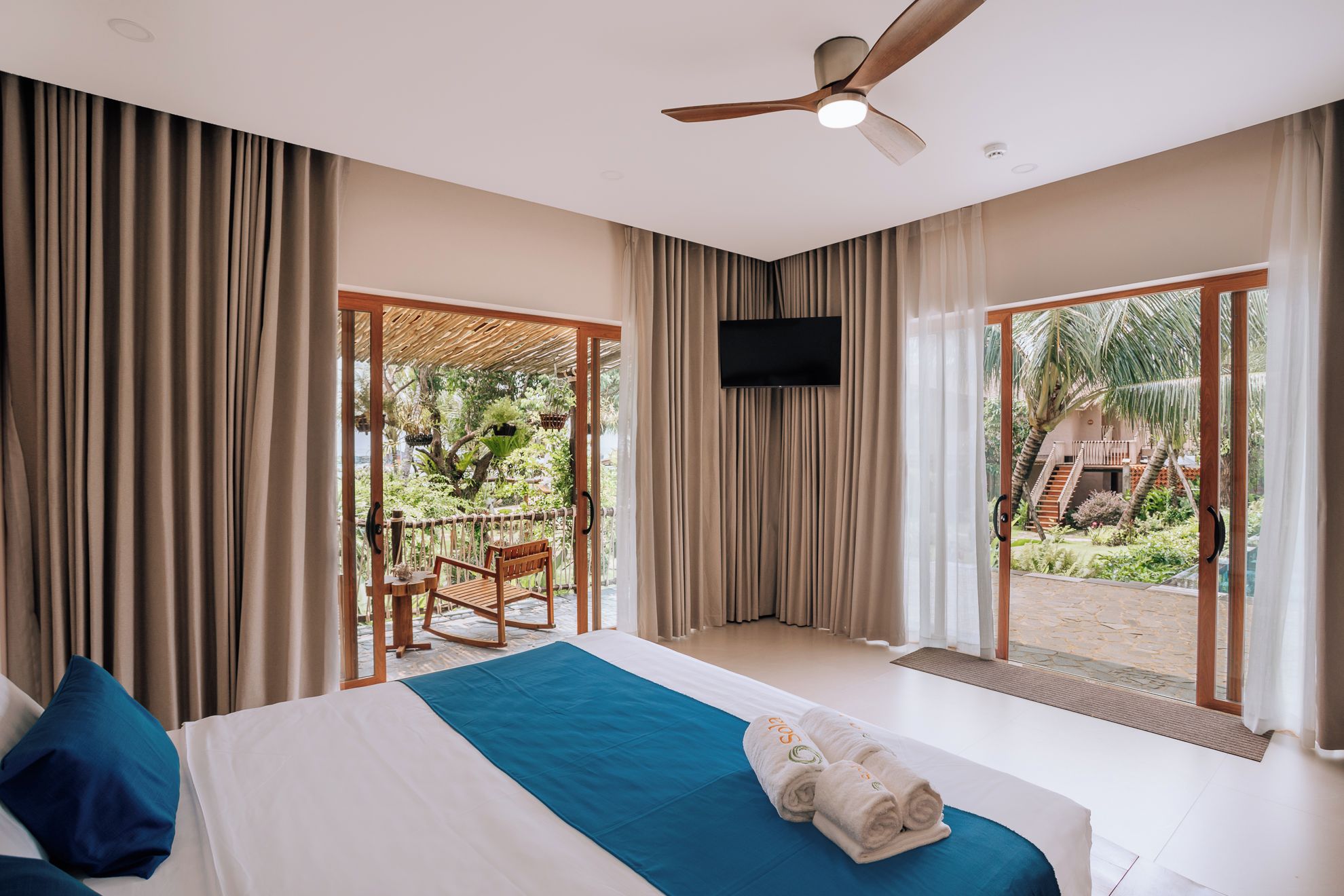 Quarto de hotel com uma cama, ventilador de teto, TV fixada na parede e vista para um jardim através de portas de vidro deslizantes.