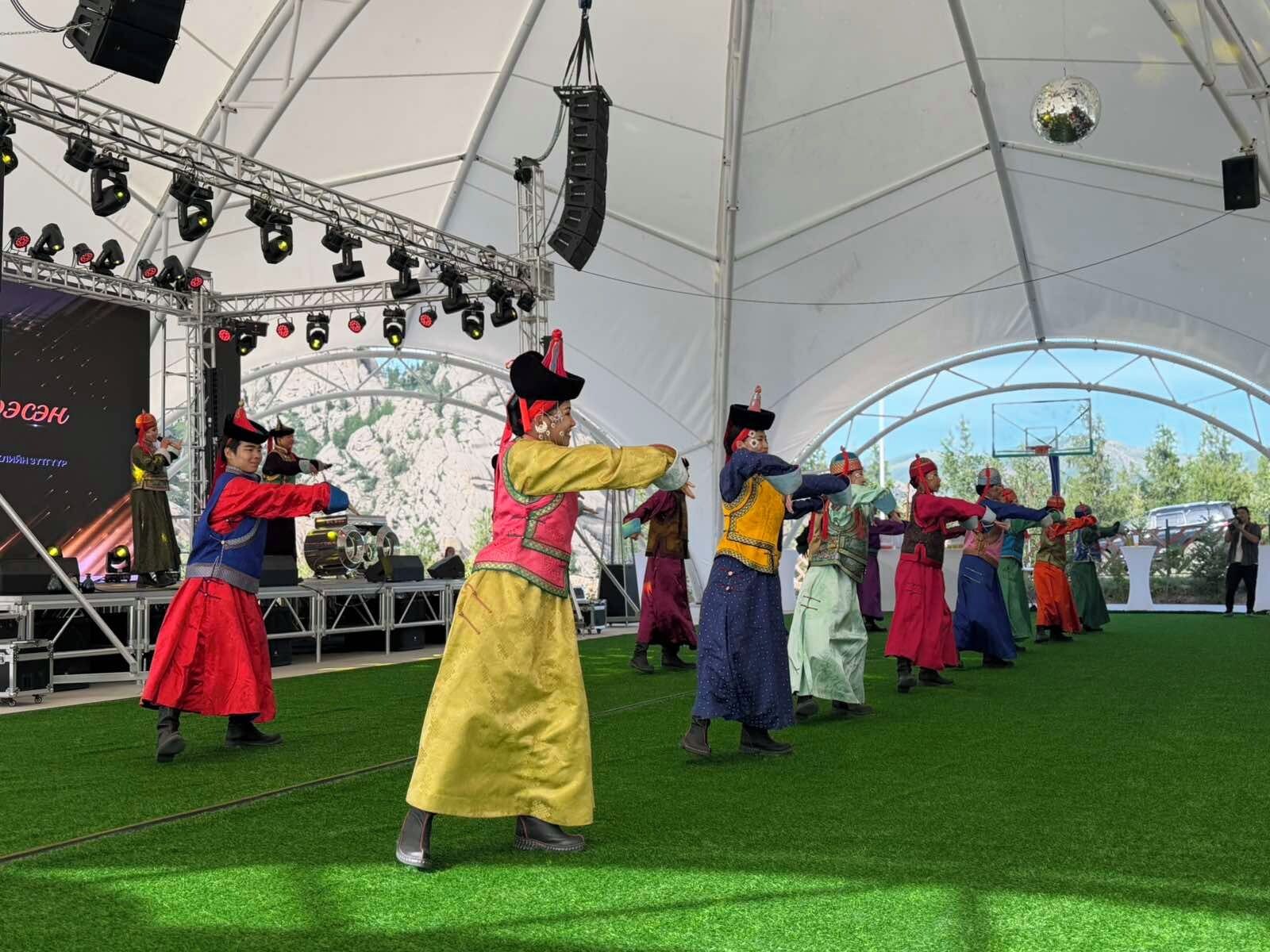 Um grupo de pessoas apresentando uma dança tradicional em um palco, com uma tenda acima e equipamento de DJ ao fundo.