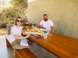 Duas pessoas sentadas à mesa de madeira, com comida e bebidas, em ambiente externo com árvores ao fundo.