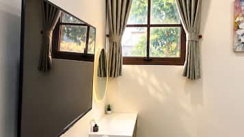 Quarto de hotel com televisão de tela plana na parede, mesa de cabeceira pequena com controle remoto, espelho e janela com cortinas.