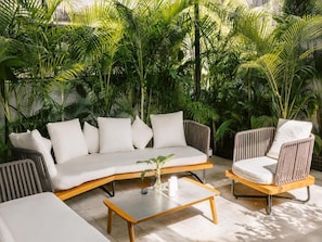 Uma área de estar moderna ao ar livre, com um sofá e poltrona, rodeada por plantas tropicais exuberantes.