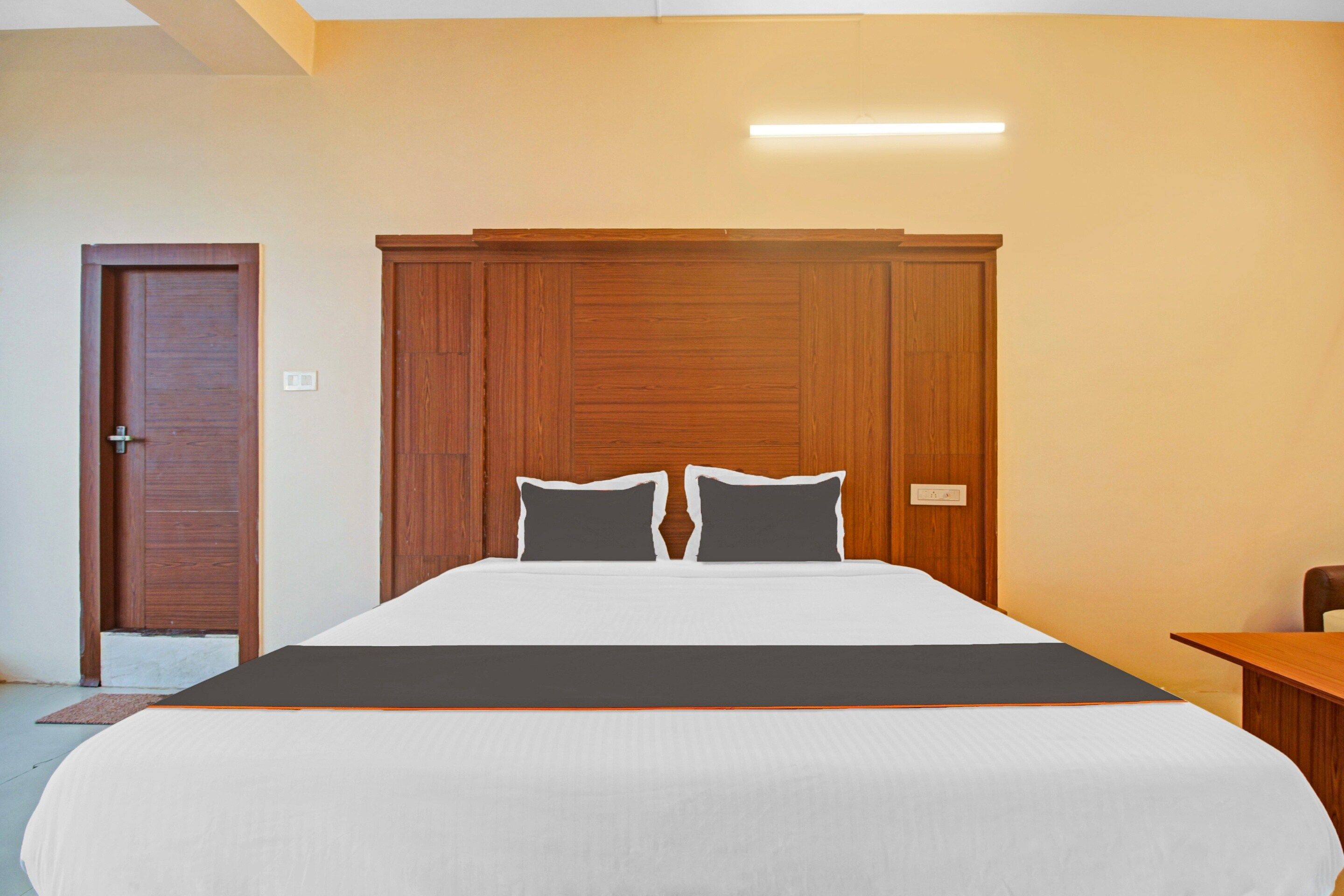 Quarto de hotel com cabeceira de madeira, cama com roupa de cama branca e duas almofadas, mesa de cabeceira e uma porta.