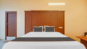 Quarto de hotel com cabeceira de madeira, cama com roupa de cama branca e duas almofadas, mesa de cabeceira e uma porta.