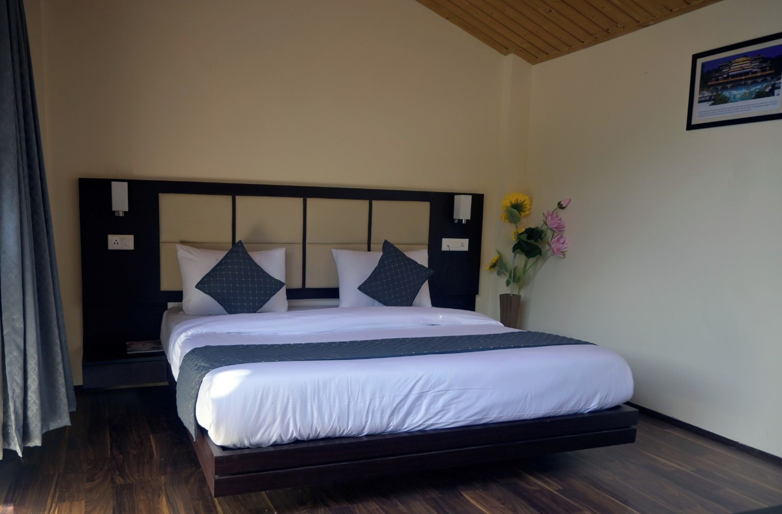 Quarto de hotel com cama, mesa de cabeceira, um vaso com flores e um quadro na parede.