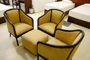 Quarto de hotel com três poltronas, um banquinho combinando, uma cama com roupa de cama branca e uma pequena mesa com abajur.