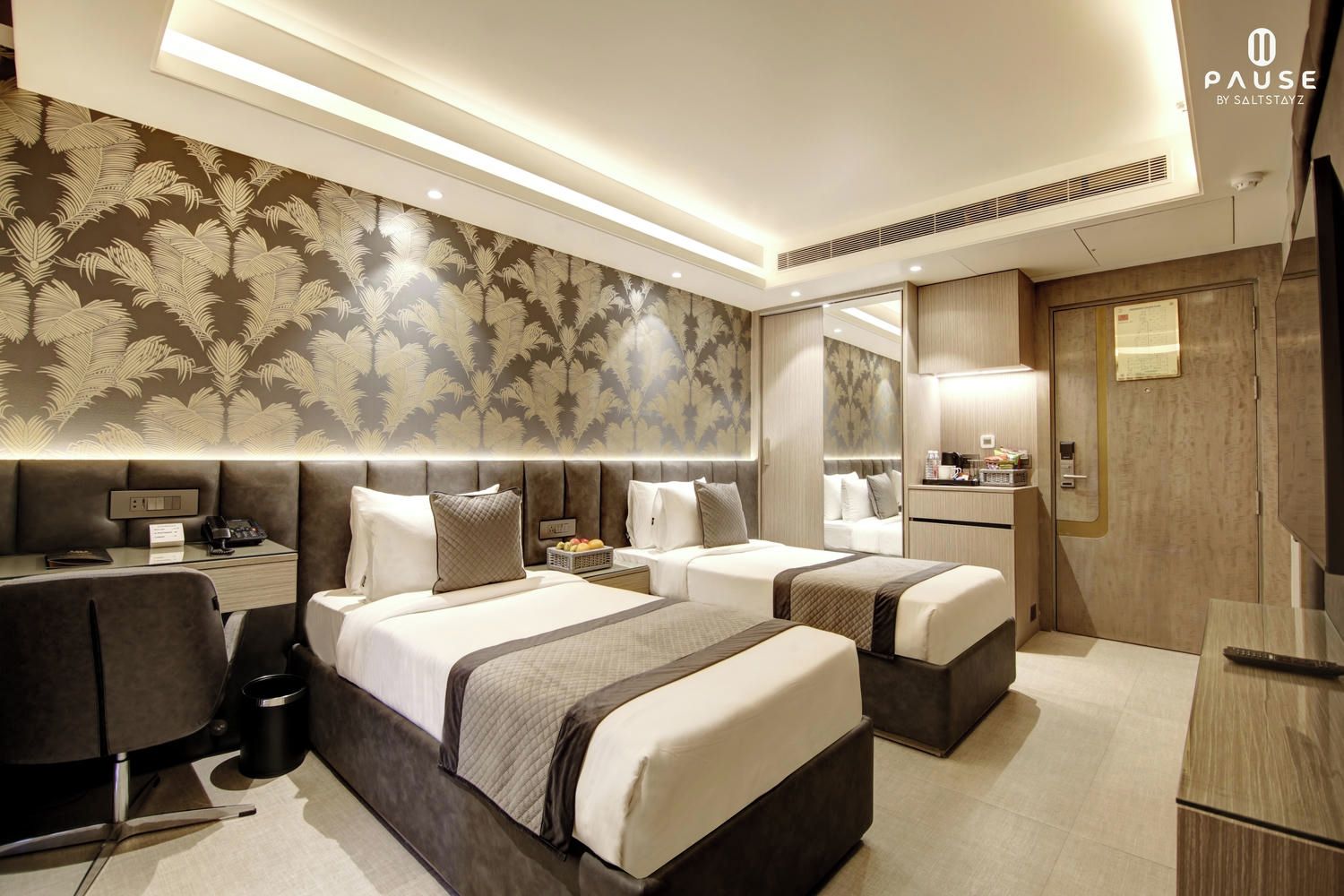 Quarto de hotel com duas camas, uma escrivaninha, uma cadeira, um cesto de lixo e um espelho.