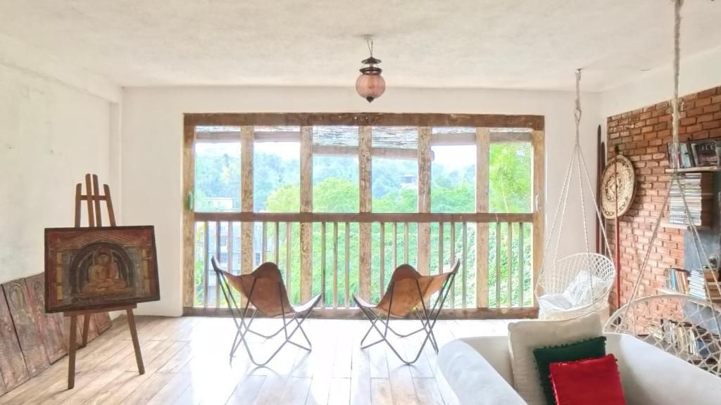 Quarto com uma grande janela, luminária pendente, cadeira branca e um quadro na parede.