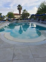 Piscina com espreguiçadeiras e guarda-sóis, rodeada por árvores e um edifício ao fundo.