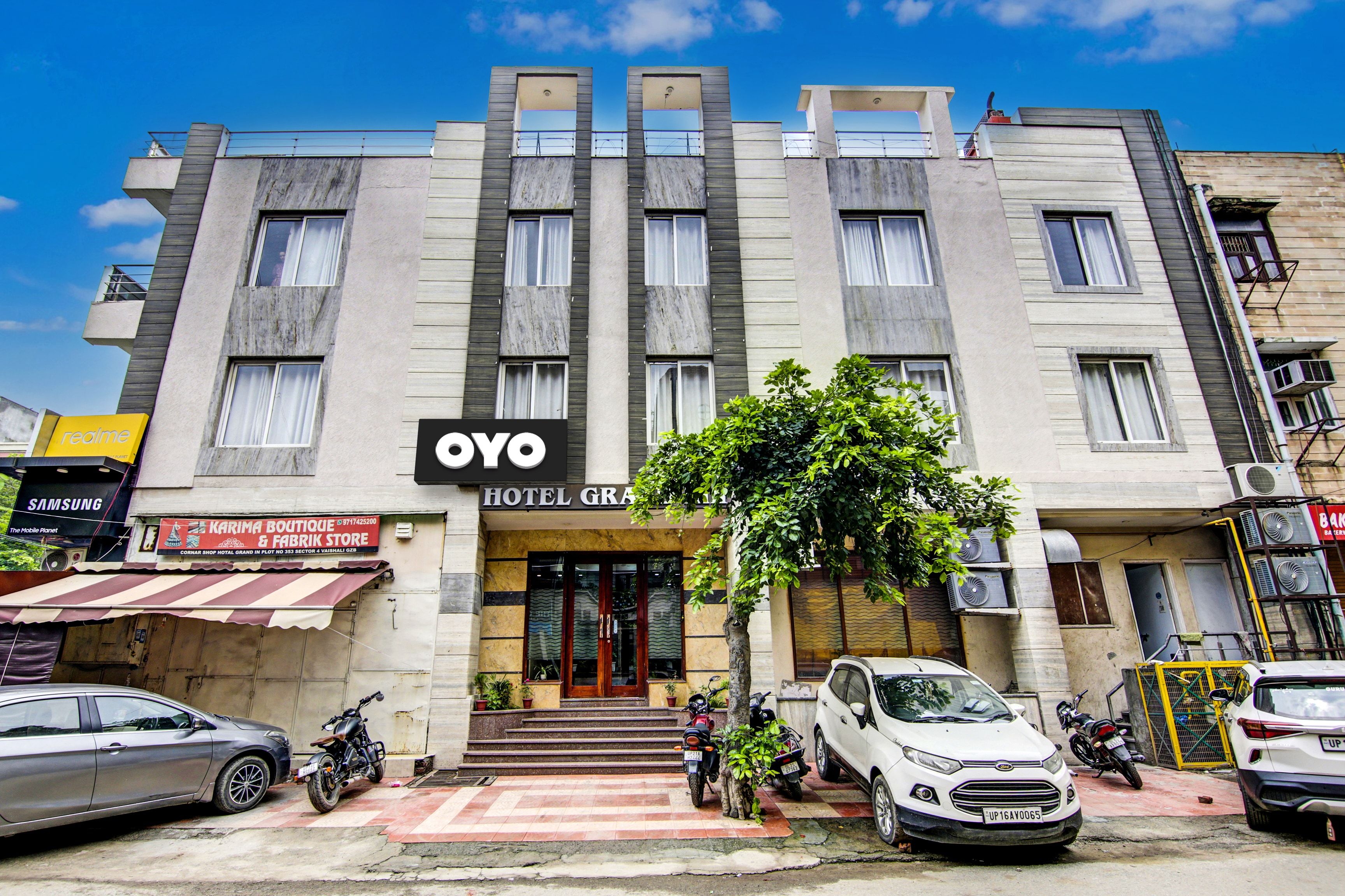 Um hotel com fachada moderna, placa indicando 'OYO HOTEL GRAND' e entrada visível com degraus até a porta.