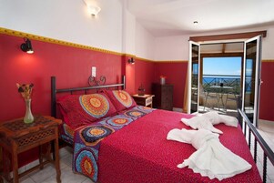 Quarto com colcha vermelha, mesa de cabeceira de madeira, vaso com flores e vista para o mar através de uma porta de varanda.