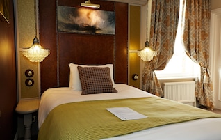 Quarto de hotel com uma cama, um quadro na parede, duas arandelas e uma janela com cortinas.