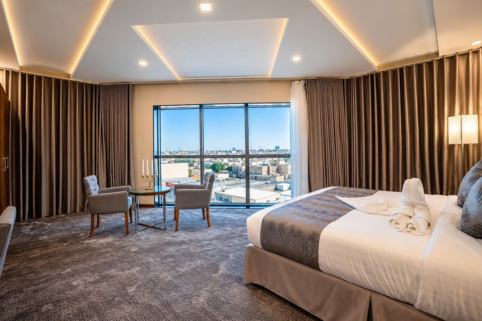 Quarto de hotel com uma cama grande, duas poltronas, uma mesa pequena e vista para a cidade através de grandes janelas.