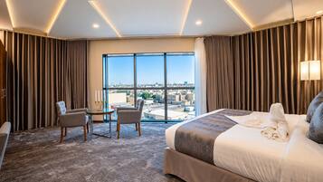Quarto de hotel com uma cama grande, duas poltronas, uma mesa pequena e vista para a cidade através de grandes janelas.