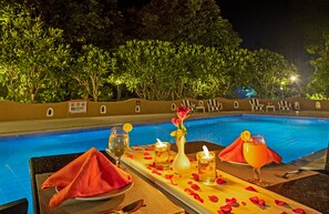 Mesa à beira da piscina com garrafa, copos e vela, cercada por vegetação e piscina.