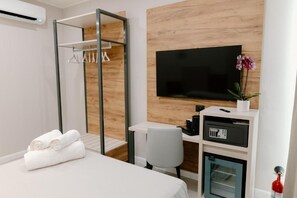 Quarto de hotel com cama, escrivaninha com televisão, mini-geladeira e closet espaçoso.