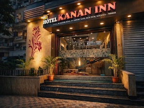 Exterior do Hotel Kanan Inn, com entrada bem iluminada, escadaria, plantas em vasos e uma placa visível.
