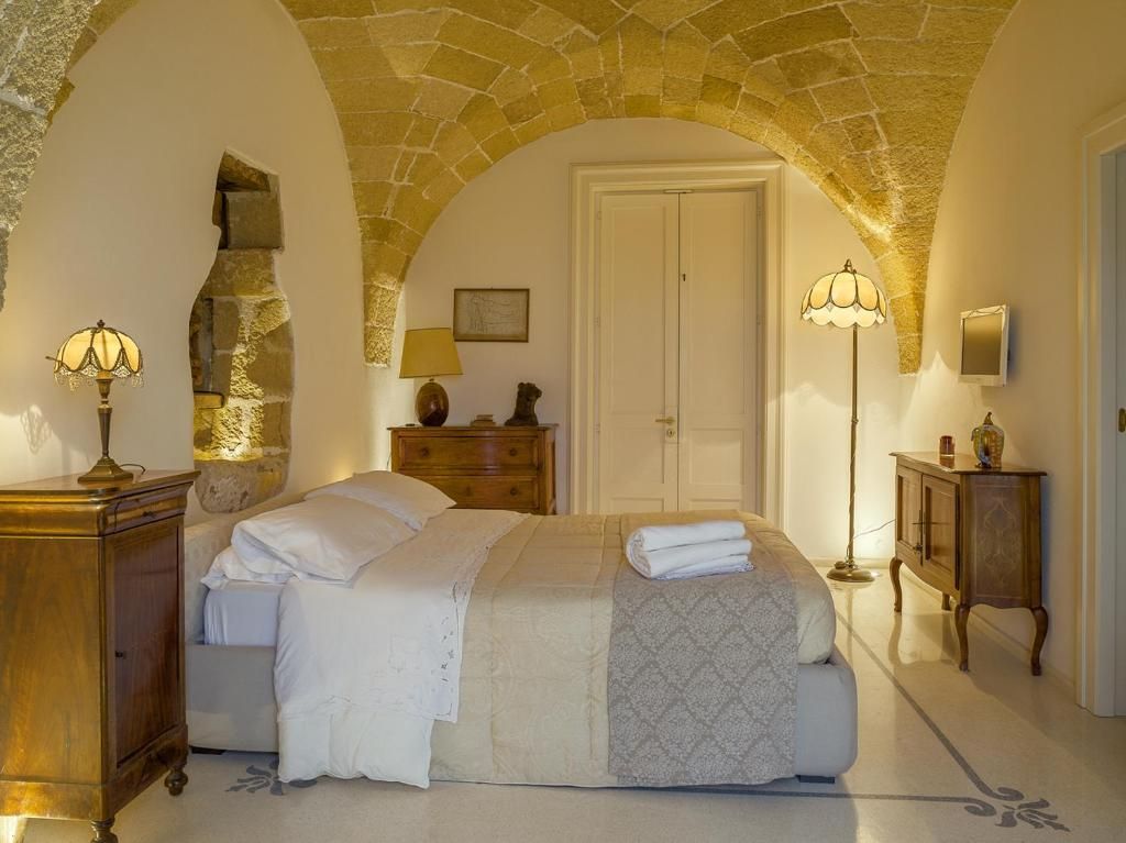 Quarto com teto em arco de pedra, cama com roupa de cama branca, cómoda de madeira e mesa de cabeceira com abajur.