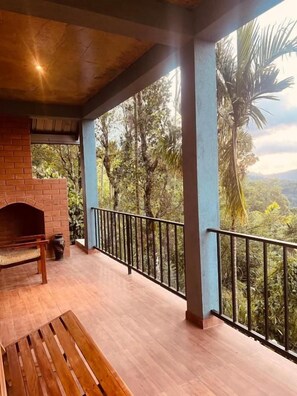 Espaço coberto ao ar livre com lareira de tijolos, banco de madeira e vista para uma paisagem arborizada.