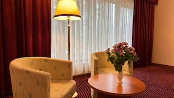 Quarto de hotel com duas poltronas, uma mesinha redonda, um vaso com flores, um abajur e uma janela com cortinas.
