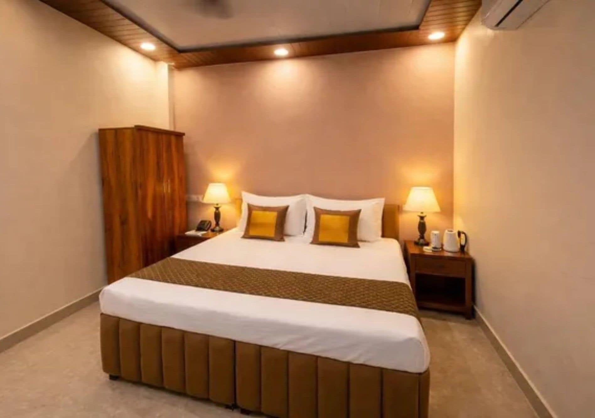 Quarto de hotel com uma cama grande, duas mesas de cabeceira com abajures, um guarda-roupa de madeira e iluminação embutida no teto.
