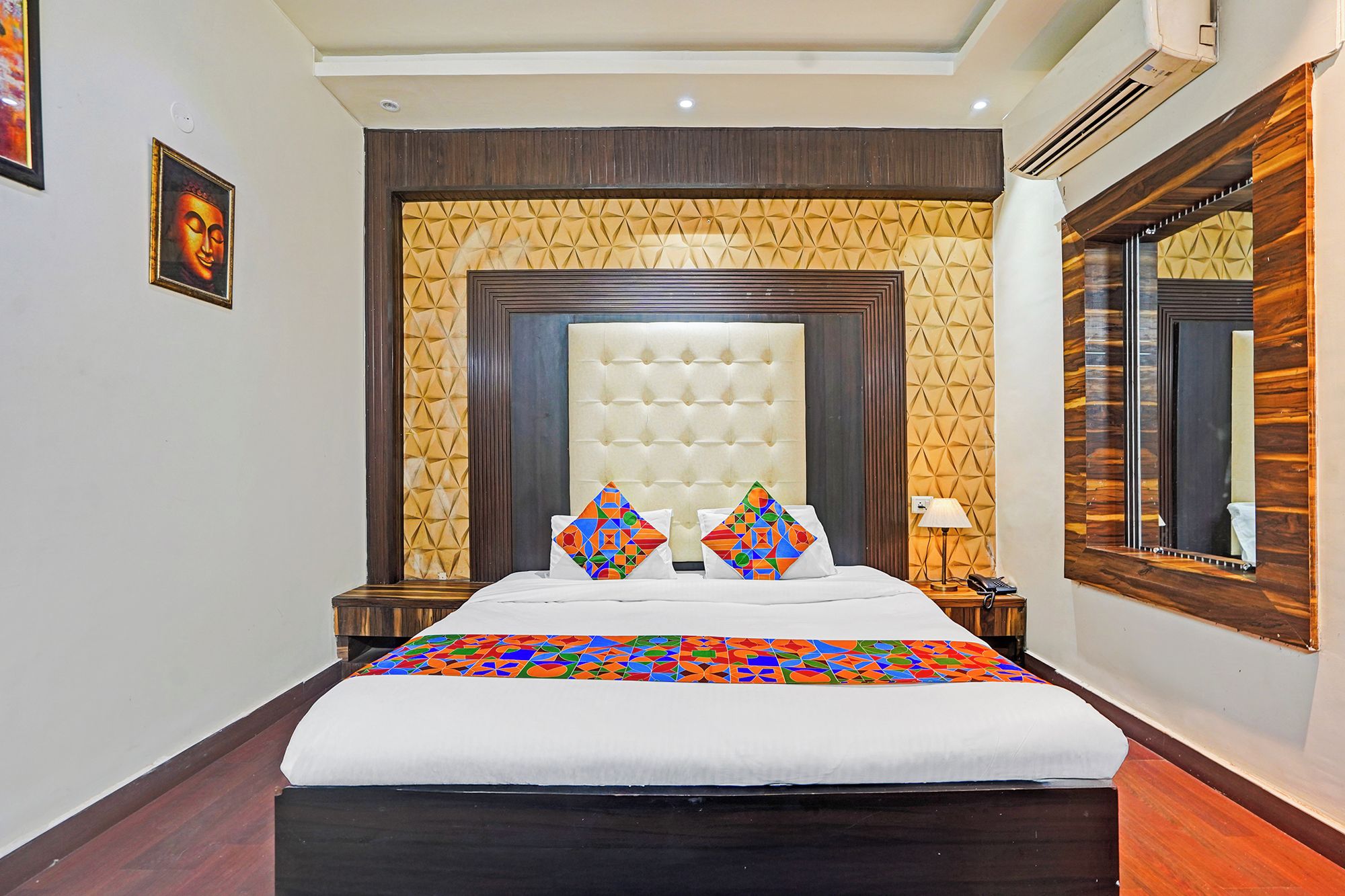 Quarto de hotel com uma cama grande, cabeceira com padrão geométrico, mesa de cabeceira com abajur e espelho na parede.