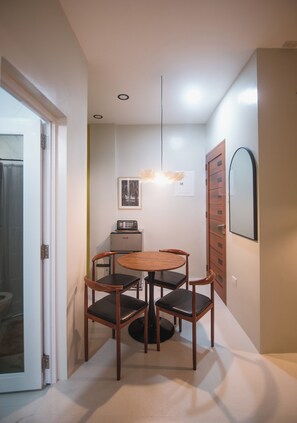 Uma área de refeições compacta com uma mesa redonda e quatro cadeiras, um frigorífico pequeno, um espelho fixado na parede e um candeeiro de teto.