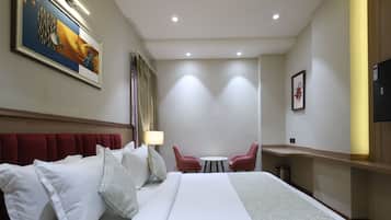 Quarto de hotel com cama, escrivaninha, duas cadeiras, abajur e um quadro na parede.
