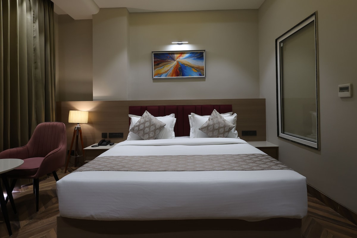 Quarto de hotel com uma cama grande, escrivaninha, cadeira, quadro na parede e uma janela ampla.