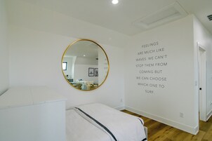 Quarto com cama, espelho redondo e parede com texto motivacional.