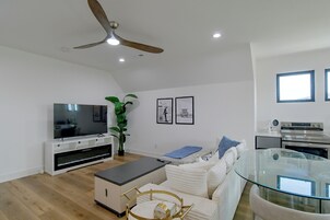 Uma sala de estar moderna com televisão de tela plana, ventilador de teto, mesa de jantar de vidro e área de cozinha com forno.