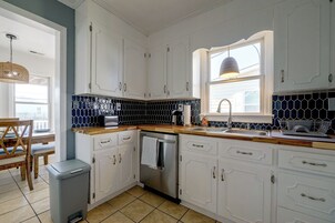 Cozinha com armários brancos, painel de azulejos pretos, bancada de madeira e janela com luminária pendente.