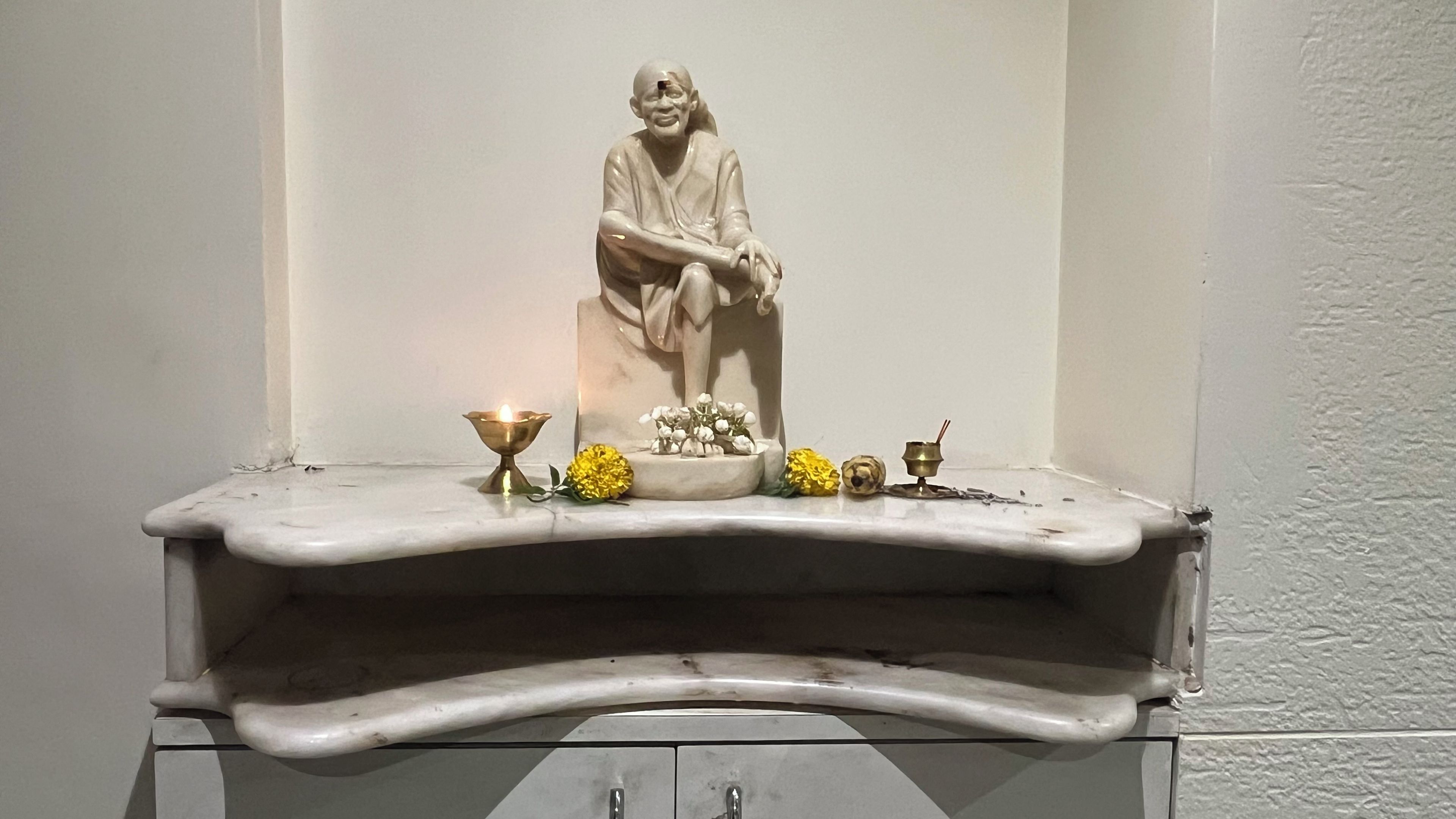 Estátua de mármore de uma figura sentada em pedestal, com uma vela acesa, flores e um objeto de latão sobre uma base de mármore branco.